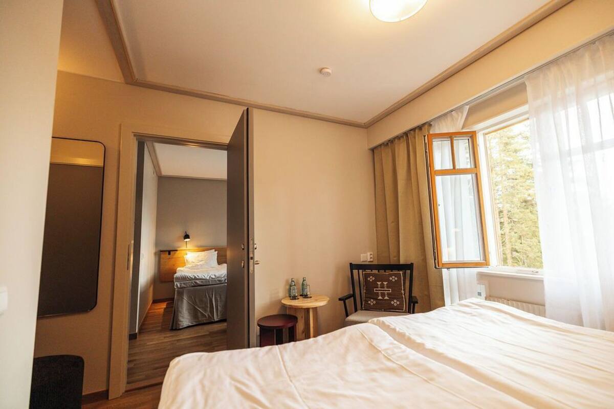 Quarto de hotel com cama, uma pequena mesa com garrafas, uma cadeira, uma janela com cortinas e uma porta que dá para outro quarto.