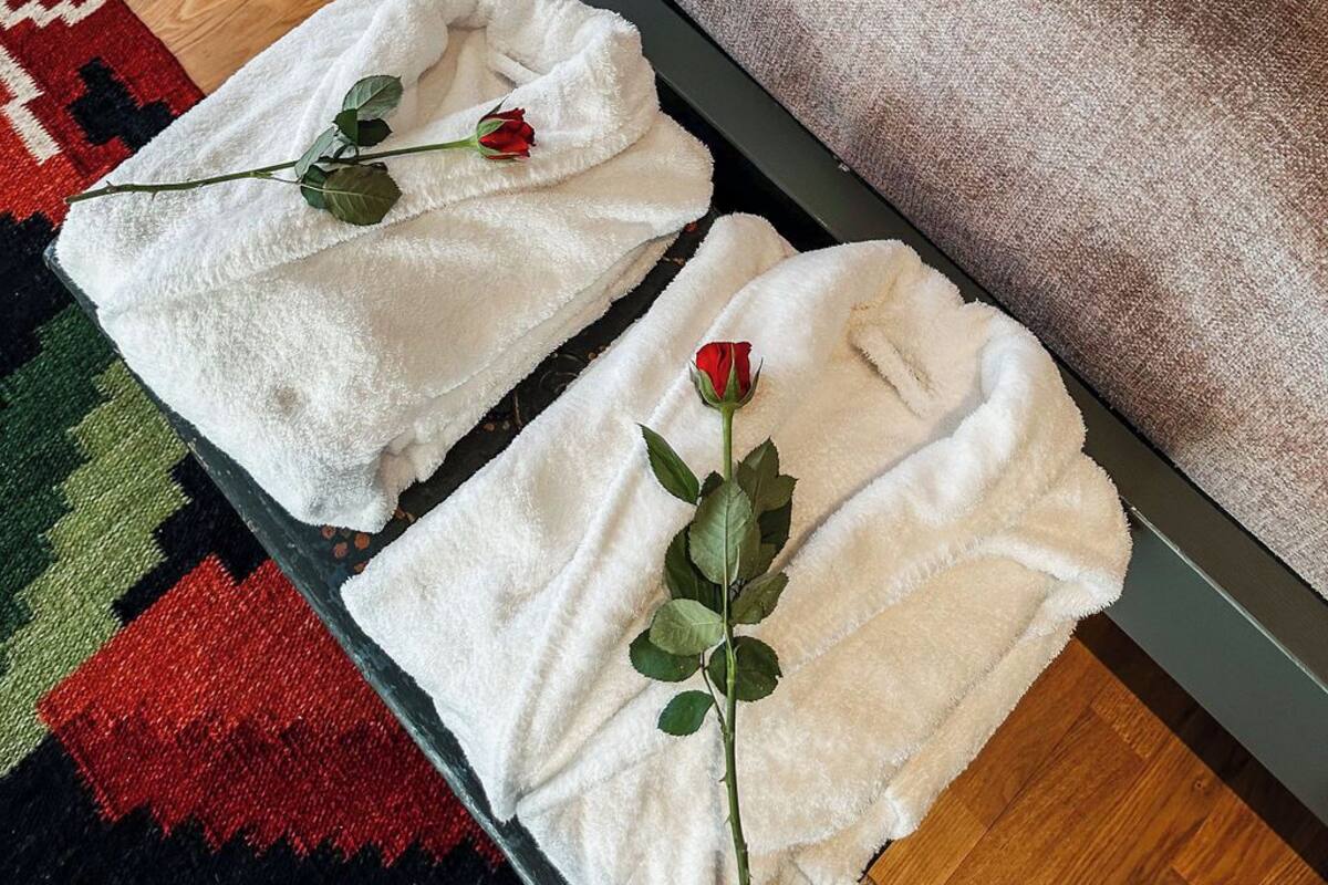 Cama com manta marrom e duas toalhas brancas com estampas de rosas sobre um piso de madeira.