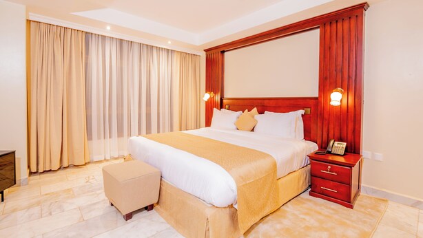 Quarto de hotel com uma cama grande, cabeceira de madeira, mesa de cabeceira com abajur e uma janela ampla com cortinas.