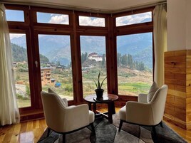 Quarto com grandes janelas que oferecem vista para uma paisagem montanhosa e um edifício tradicional. Há duas cadeiras e uma pequena mesa com uma planta.