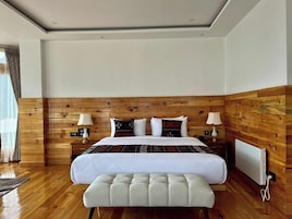 Quarto com cabeceira de madeira, uma cama grande com roupa de cama branca, duas mesas de cabeceira com abajures e um banco branco.