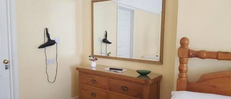 Quarto com cômoda de madeira, espelho, cama com cabeceira de madeira e secador de cabelo fixado na parede.