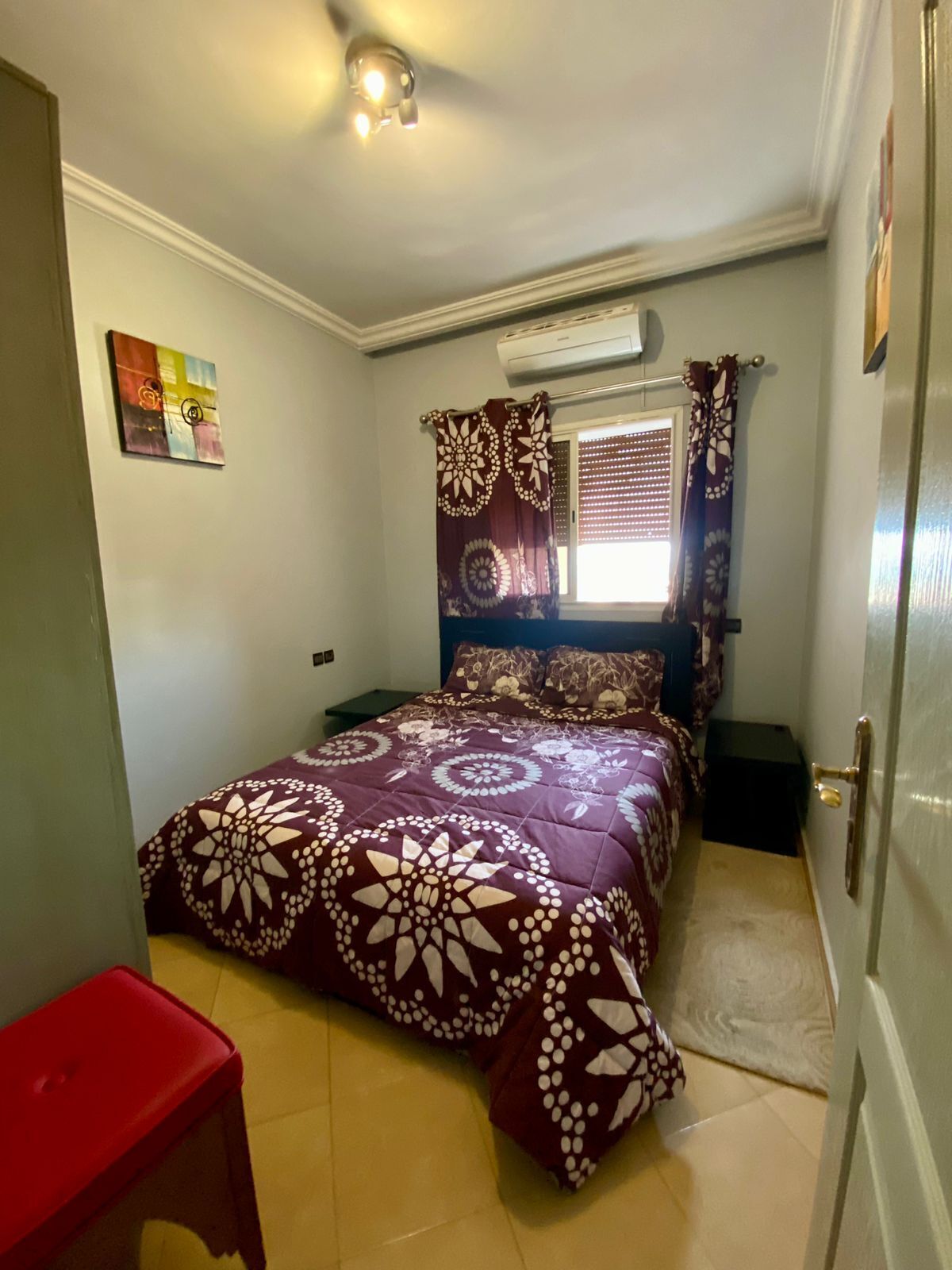 Quarto com cama, ar condicionado, janela com cortinas, uma cadeira vermelha e uma pequena mesa.