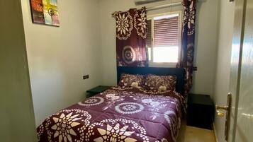 Quarto com cama, ar condicionado, janela com cortinas, uma cadeira vermelha e uma pequena mesa.