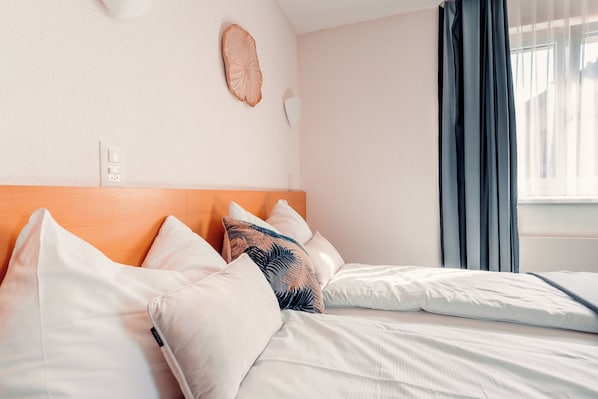 Cama bem arrumada com roupa de cama branca e cabeceira de madeira, luminária fixada na parede e janela com cortinas.