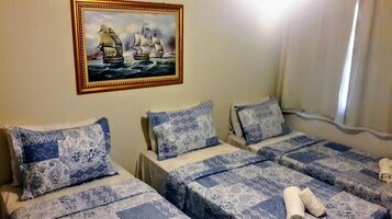 Quarto com três camas, cada uma com roupa de cama estampada em azul e branco. Um quadro de um navio à vela está pendurado na parede.