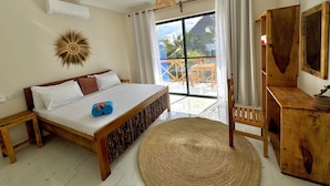 Quarto com cama, ventoinha no teto, uma janela grande com cortinas, uma cadeira de madeira e uma mesinha.