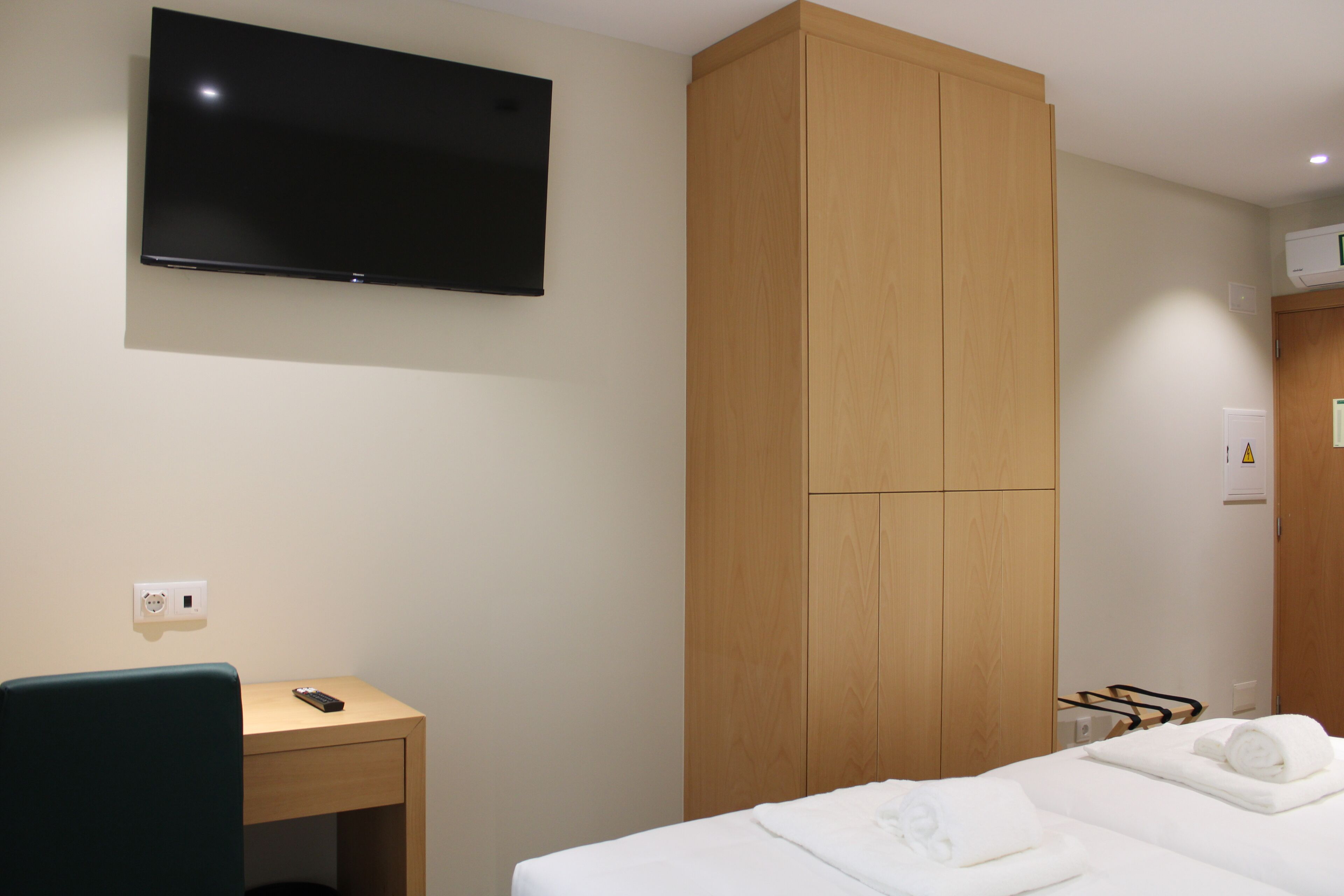 Quarto de hotel com uma cama, uma escrivaninha, televisão e guarda-roupa.