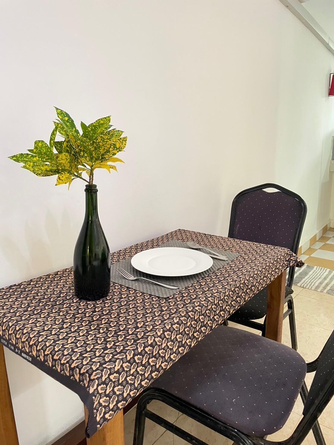 Uma mesa de jantar com toalha de mesa estampada, um prato branco, talheres e uma planta verde num vaso.