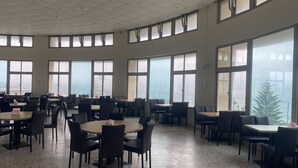 Uma sala de jantar ampla, com várias mesas e cadeiras, janelas grandes e teto alto.