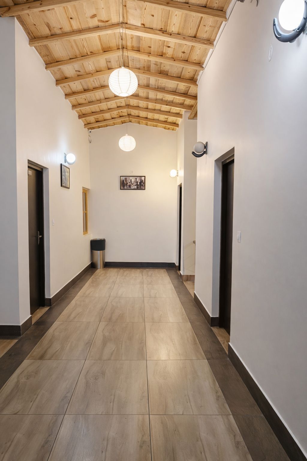 Um corredor com piso de madeira, paredes brancas e teto de madeira. Há duas portas de cada lado, uma lixeira e um quadro na parede.