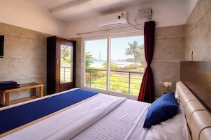 Quarto de hotel com cama, uma pequena mesa, televisão e janela com vista para a praia e palmeiras.
