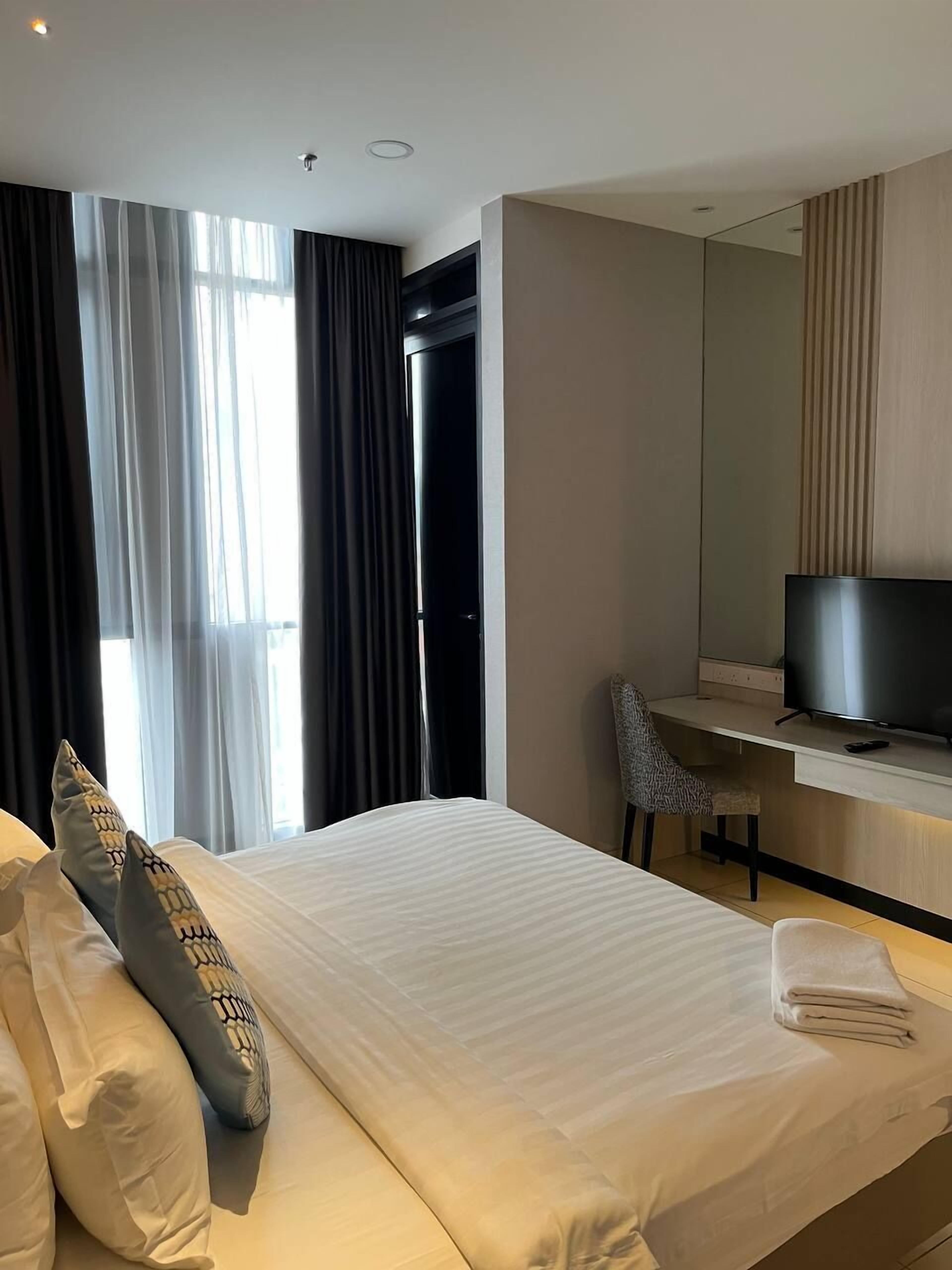 Quarto de hotel com cama, escrivaninha com televisão, uma cadeira e uma janela grande com cortinas.
