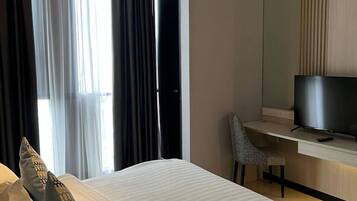 Quarto de hotel com cama, escrivaninha com televisão, uma cadeira e uma janela grande com cortinas.