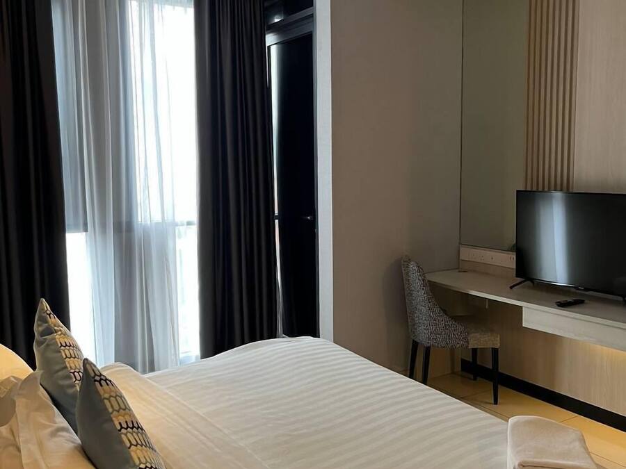 Quarto de hotel com cama, escrivaninha com televisão, uma cadeira e uma janela grande com cortinas.