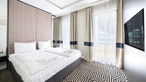 Quarto de hotel com uma cama grande, cabeceira de madeira, televisão de tela plana e vista para um edifício através de cortinas translúcidas.