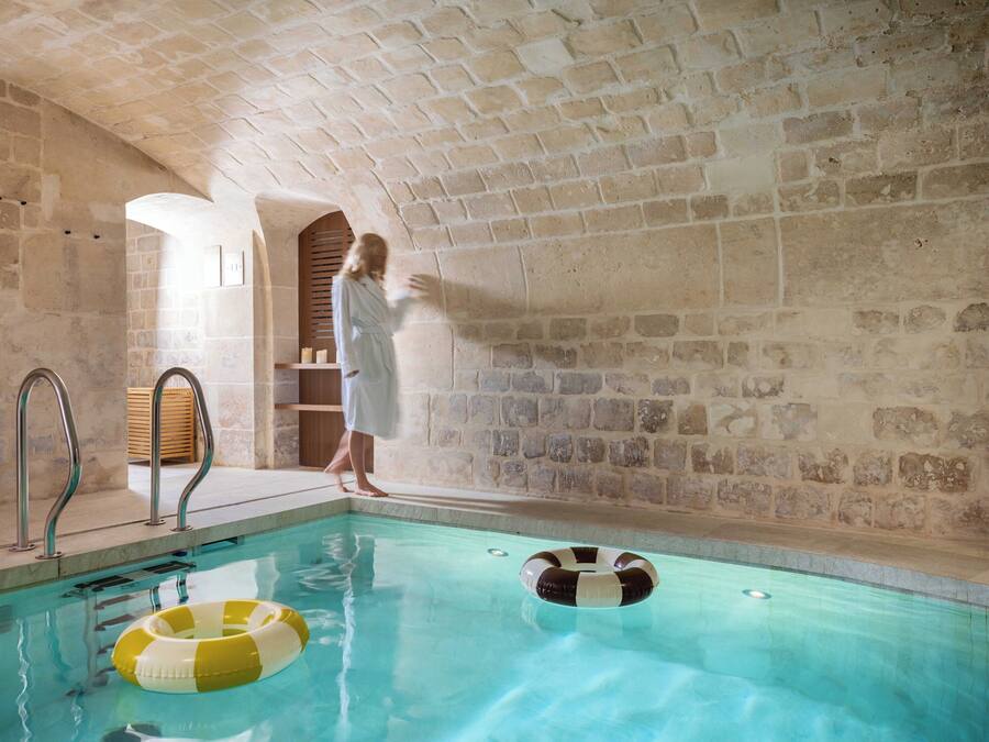 Uma piscina interior com paredes de pedra, água cristalina, uma escada de metal e dois flutuadores infláveis.