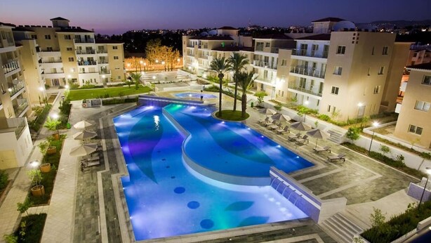 Piscina rodeada por edifícios modernos com varandas, área ajardinada com palmeiras e mobiliário para áreas externas.