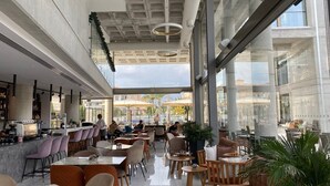 Uma área de refeições interna moderna, com pé-direito alto, grandes janelas e diversas opções de assento, incluindo mesas e cadeiras.