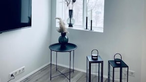 Quarto moderno com uma grande janela, televisão de tela plana na parede, uma mesa redonda com um vaso de plantas secas e duas mesinhas laterais pretas.