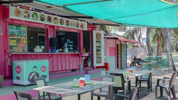 Um carrinho de comida com fachada rosa, balcão de autoatendimento e uma geladeira exibindo bebidas. Cadeiras e mesas de plástico do lado de fora.