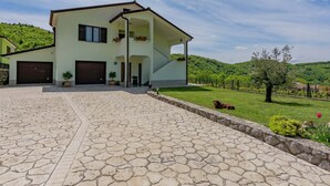 Uma casa moderna de dois andares, com um caminho de entrada pavimentado, varanda coberta e vista para uma encosta verde.