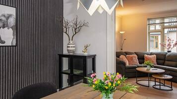 Uma sala de estar moderna com uma mesa de jantar de madeira, uma mesinha lateral preta, um sofá com almofadas, um abajur e um vaso com flores.