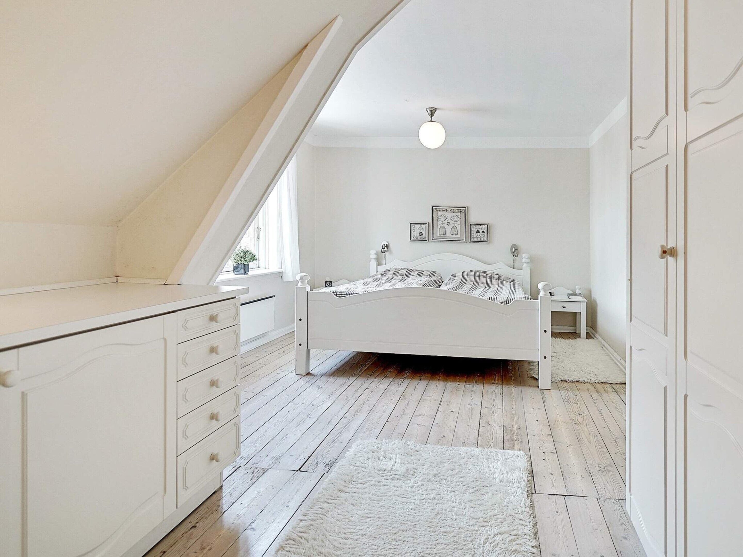 Um quarto com teto inclinado, uma cama com cabeceira branca, cômoda com gavetas, mesa de cabeceira pequena e um ventilador de teto.