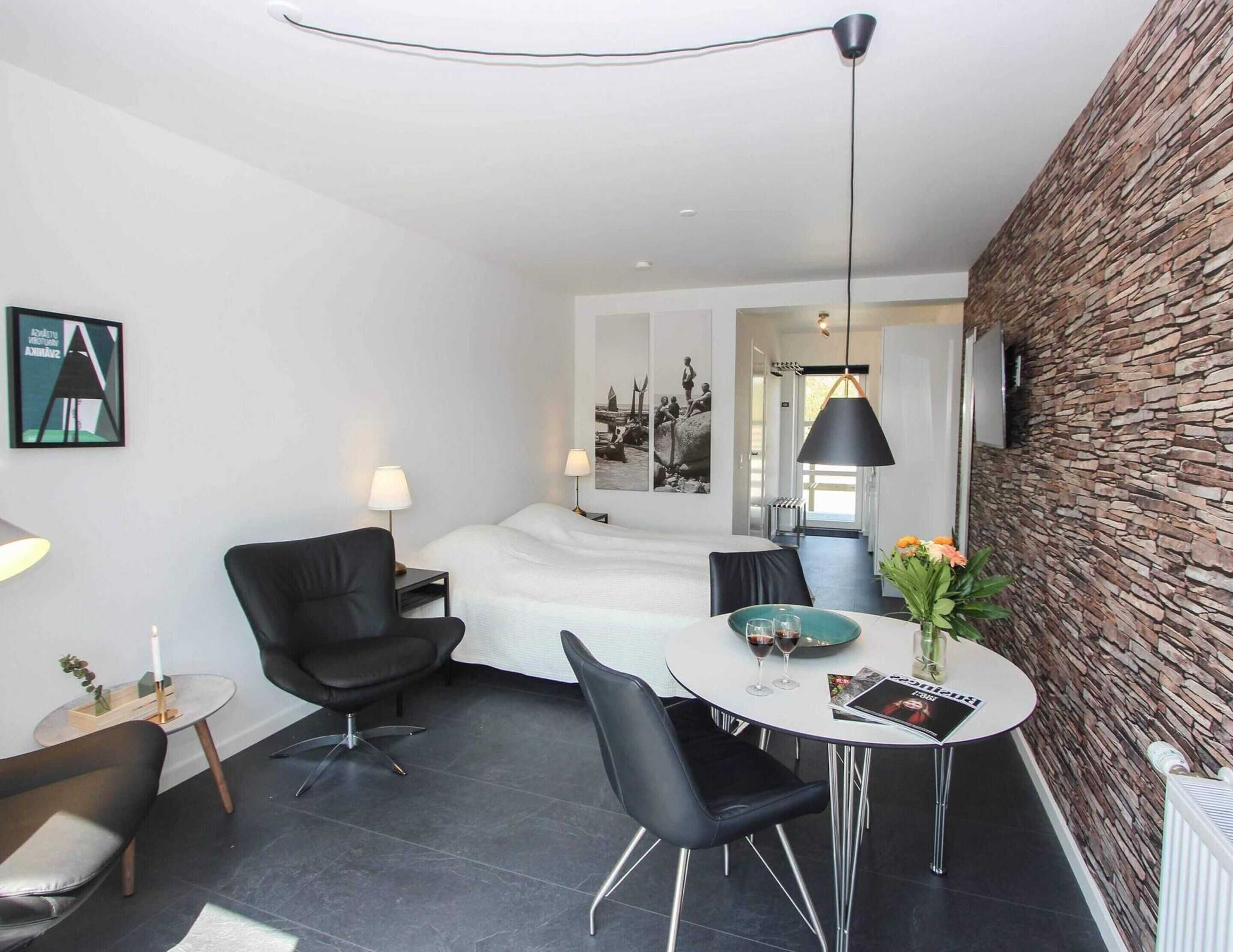 Quarto de hotel moderno com cama, mesa de jantar com cadeiras, poltrona de couro preta, mesinha lateral com luminária e parede de pedra.