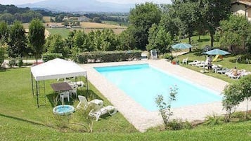 Piscina retangular com uma tenda branca e cadeiras de descanso em área gramada, cercada por árvores e vista para montanhas distantes.