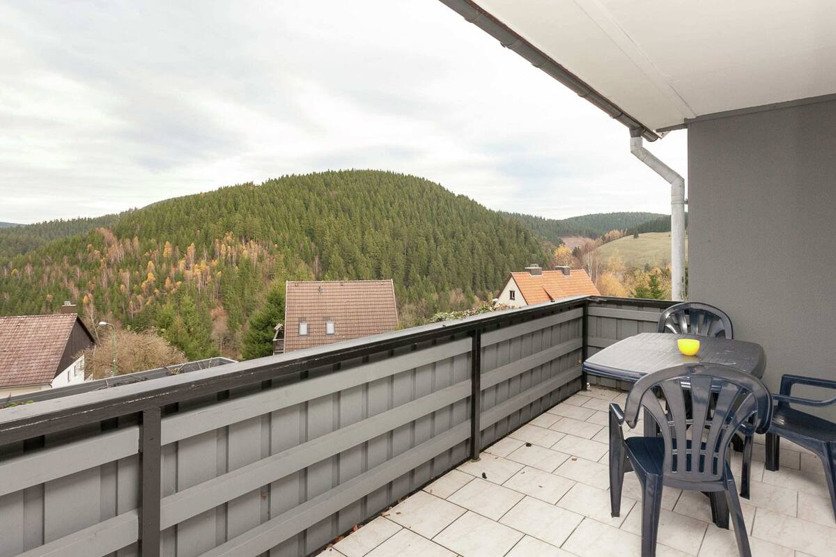Varanda com vista para uma colina arborizada, mesa com cadeiras e chão de azulejos brancos.