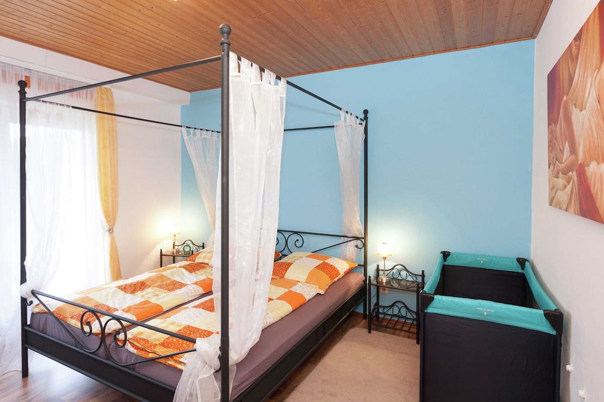 Uma cama com dossel e cortina, uma mesa de cabeceira com abajur, e uma pequena prateleira com almofada azul-turquesa.