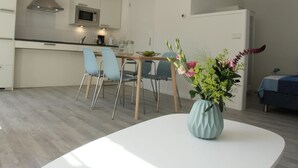 Cozinha moderna com armários brancos, micro-ondas e exaustor de aço inoxidável. Há uma área de refeições com mesa branca e cadeiras azuis. Um vaso com flores está sobre a mesa.