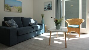 Uma sala de estar moderna com um sofá azul escuro, uma poltrona de vime, uma mesa redonda pequena e duas pinturas penduradas na parede.