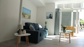 Uma sala de estar moderna com um sofá azul escuro, uma mesinha redonda e uma cadeira de vime. Há um quadro na parede e uma janela com cortinas.