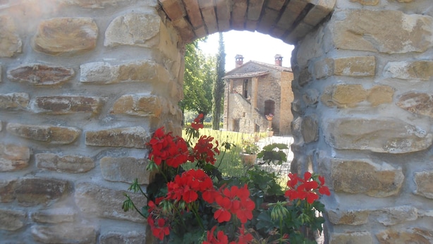Um arco de pedra com flores vermelhas em primeiro plano e um edifício com telhado de azulejos ao fundo.