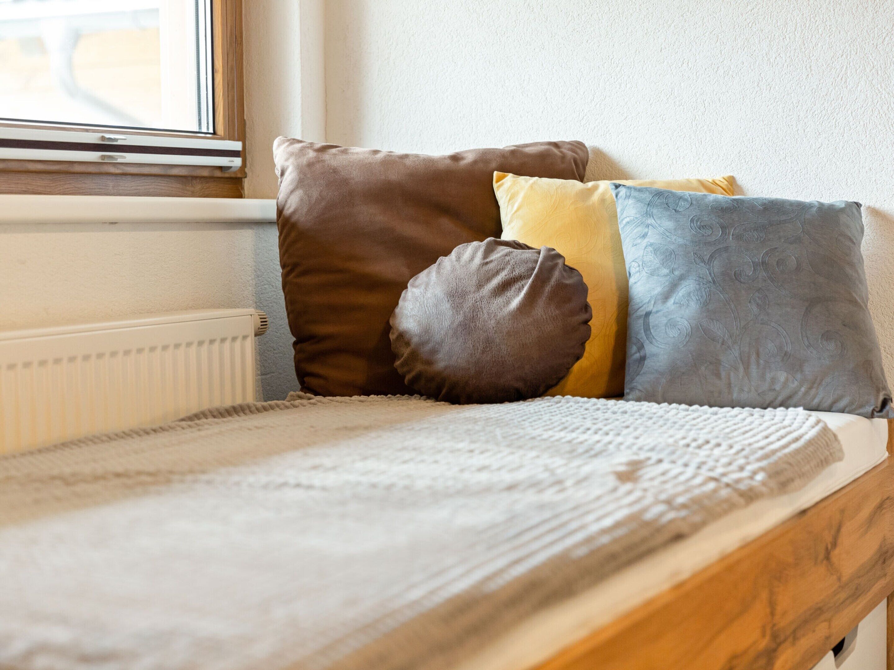 Cama com colchão branco e manta texturizada, adornada por quatro almofadas em cores e padrões variados.