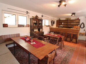 Uma área de refeições acolhedora com uma mesa de madeira, cadeiras e um tapete vermelho. Há também um balcão com banquetas e prateleiras com potes e garrafas.