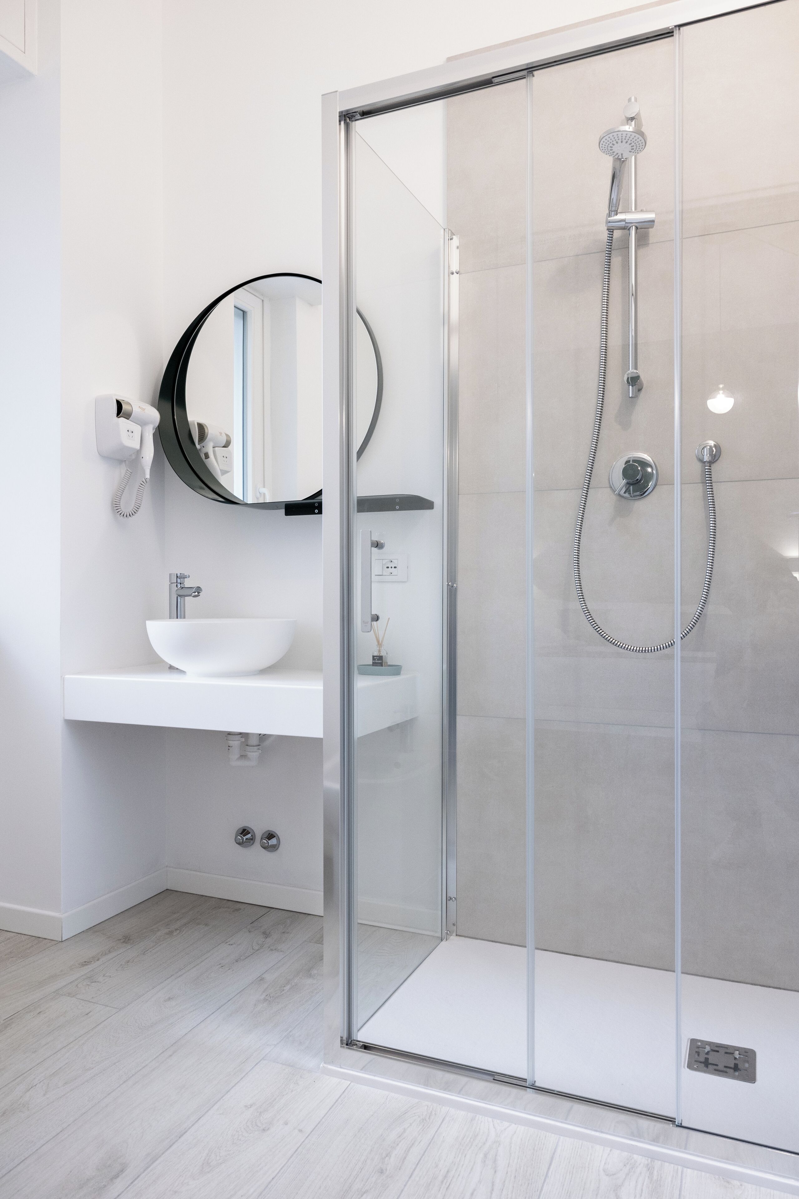 Um banheiro moderno com chuveiro em box de vidro, pia branca com espelho redondo e secador de cabelo fixado na parede.
