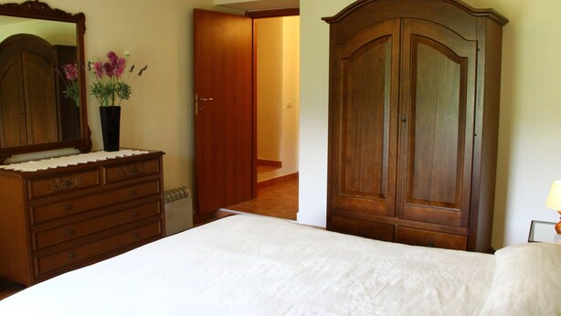Quarto com cômoda de madeira, espelho, cama e porta para outro aposento.