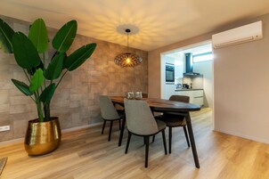Uma sala de jantar moderna com mesa de madeira, quatro cadeiras, uma planta verde grande em vaso dourado e uma luminária de teto singular.