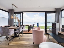 Uma área de refeições moderna com vista para o mar, apresentando uma mesa de madeira, cadeiras cinzentas e rosa, e uma televisão sobre um móvel.