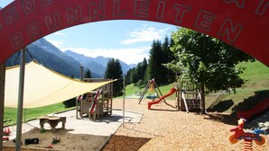 Um parquinho com um arco vermelho com a inscrição "UNDERSPIELPLAT BRUNNLEITEN" e diversos equipamentos de recreação.