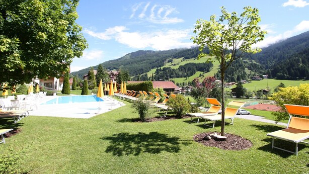 Área de piscina com espreguiçadeiras e uma pequena árvore, cercada por um gramado e montanhas ao fundo.