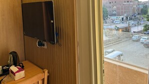 Quarto com parede de painel de madeira, televisão de tela plana montada, pequena mesa de madeira com chaleira e caixa de lenços, e vista para uma rua movimentada e edifícios lá fora.