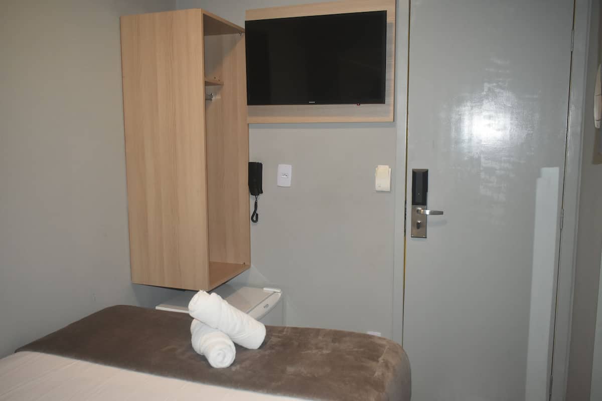 Um quarto pequeno com uma cama, um armário de madeira, uma televisão e uma porta.