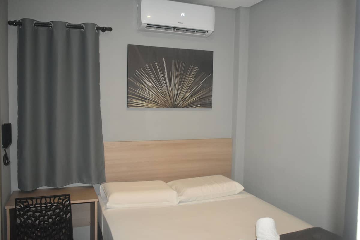 Quarto com cama, cabeceira, ar condicionado fixado na parede, cortina e uma obra de arte na parede.