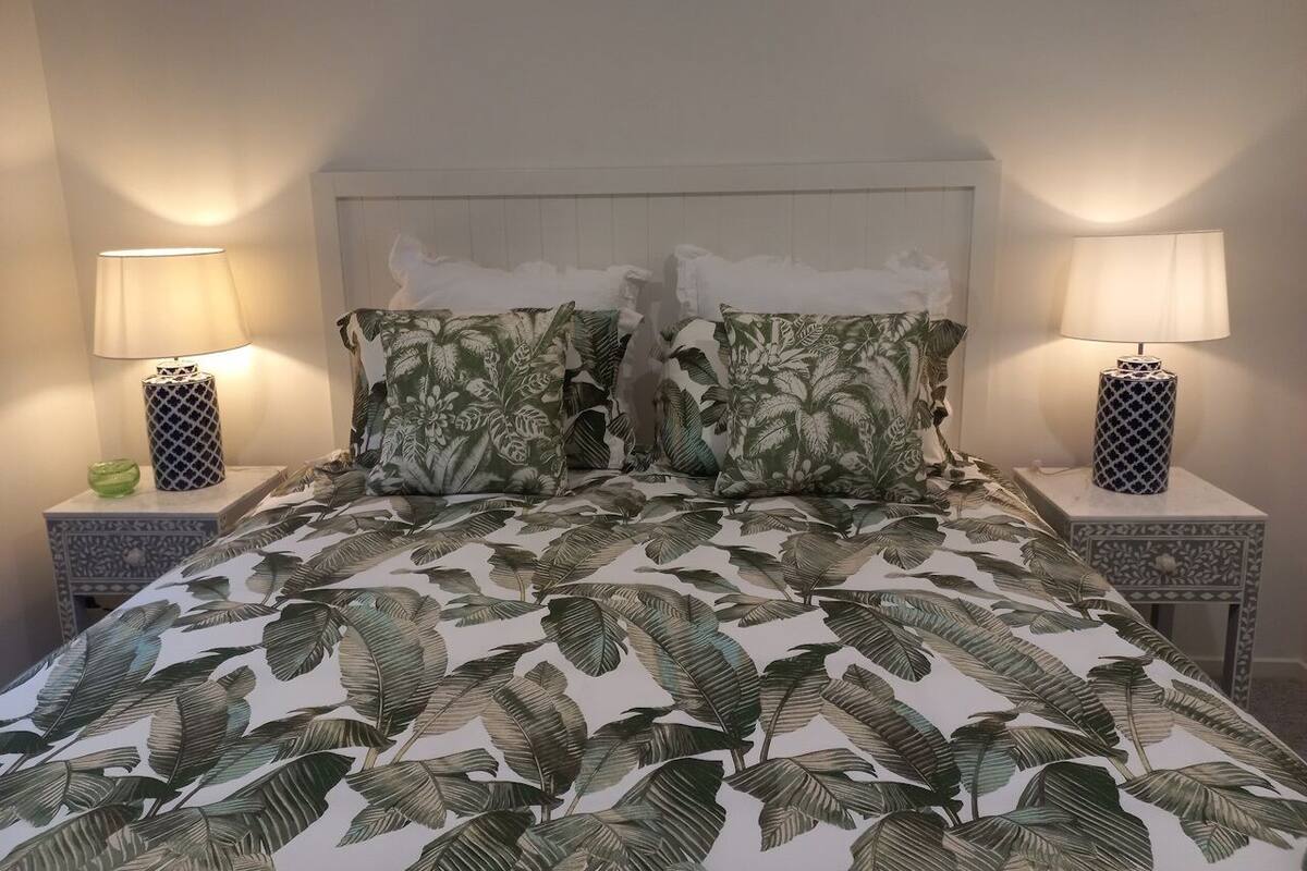Uma cama com colcha estampada em folhagens, duas almofadas iguais e duas mesas de cabeceira com abajures e vasos decorativos.