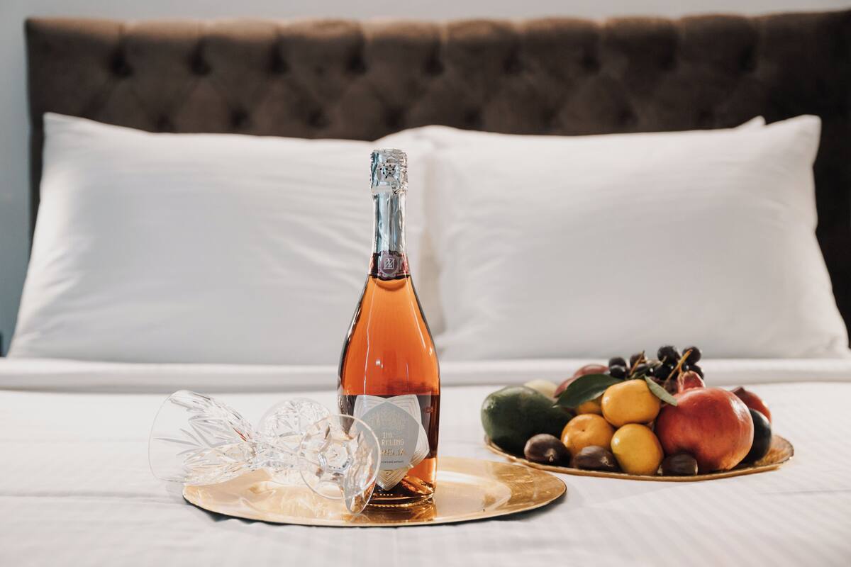 Uma cama com cabeceira estofada, uma garrafa de champanhe, um copo e um prato de frutas sobre um lençol branco.