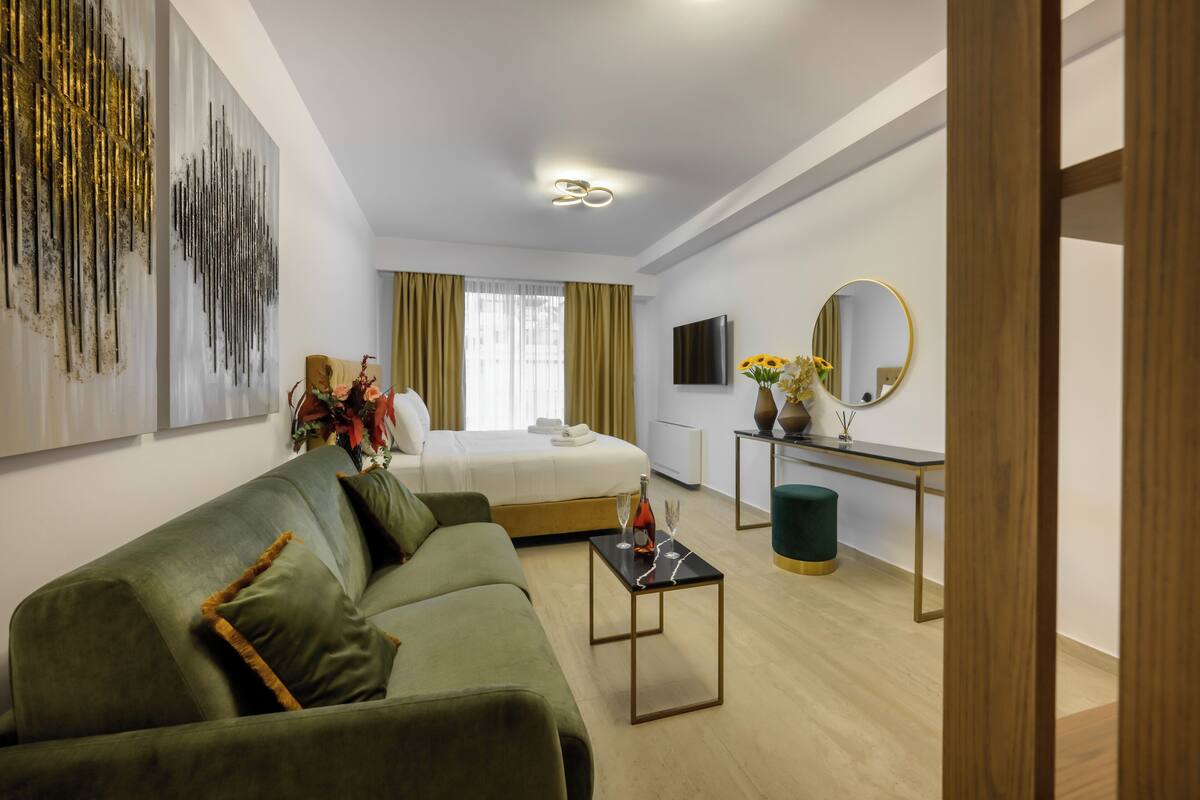 Quarto de hotel moderno com uma cama grande, um sofá verde, uma mesinha com uma garrafa e copos, uma televisão e um espelho.
